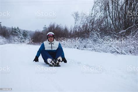 푸른 옷을 입은 젊고 행복한 남성 스노우보더가 언덕 아래로 미끄러지면서 스노우 보드에 앉아 있습니다 겨울에 대한 스톡 사진 및 기타 이미지 Istock