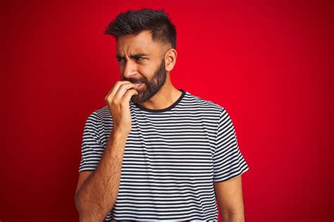 젊은 잘 생긴 인도 남자 입고 해군 줄무늬 티셔츠 위에 고립 된 빨간 배경 찾고 스트레스와 긴장 에 손을 물고 손톱 불안 문제 T 셔츠에 대한 스톡 사진 및 기타 이미지