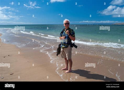 Smiling Barefoot Man With A Camera Standing In Ocean Surf On A Beach