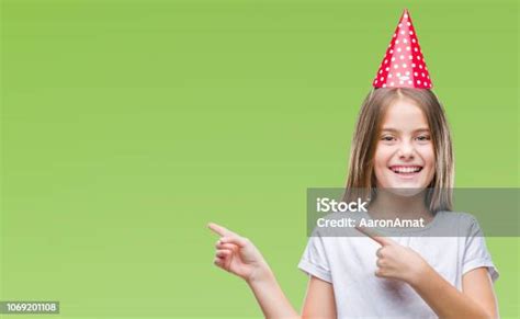 젊은 아름 다운 소녀를 통해 생일 모자를 쓰고 웃 고 옆에 두 손과 손가락으로 가리키는 카메라를 보고 배경 고립 아이에 대한 스톡 사진 및 기타 이미지 Istock