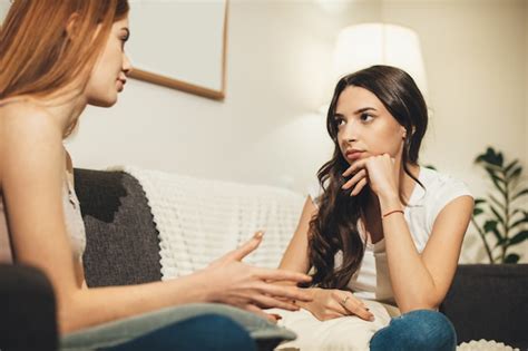 Señora caucásica de jengibre con pecas sentada en el sofá con su amiga morena y discutiendo