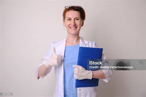 하얀 코트를 입은 젊은 빨간 머리의 여성 메딕은 손에 메모를 하고 미소를 짓는 폴더를 가지고 있습니다 그녀는 엄지 손가락을 보여주는 35 39세에 대한 스톡 사진 및 기타