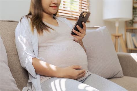 Happy Asian Pregnant Woman Using Smart Phone While Relaxing On Sofa In