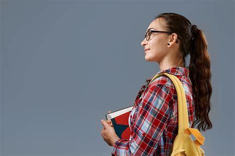 안경 제곱 셔츠를 입은 젊은 여성에게는 노란색 배낭과 큰 책이 있습니다 회색 스튜디오 배경에 서 있는 학생 여성의 초상화 얼간이에 대한 스톡 사진 및 기타 이미지 Istock