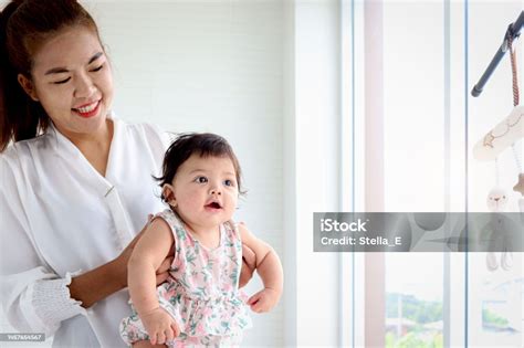행복한 미소를 짓고 있는 사랑스러운 6개월 아기 소녀는 엄마의 팔을 껴안고 창문 옆에 서 있고 엄마는 귀여운 어린 딸 아이를