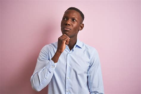 アフリカ系アメリカ人男性は孤立したピンクの背景の上に立つ青いエレガントなシャツを着て質問ペンシブな表情を考えてあごに手を置いて思慮深い顔で