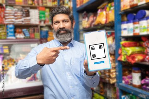 Portrait Of Happy Mature Indian Man Showing Qr Code Scanner To Pay Online At Grocery Shop Or