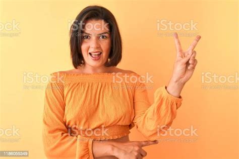 고립 된 오렌지 색 배경 위에 서있는 캐주얼 한 티셔츠를 입은 젊은 아름다운 여성은 승리의 표지판을하고있는 카메라에서 행복한 얼굴 윙크와 미소 2번 T 셔츠에 대한 스톡