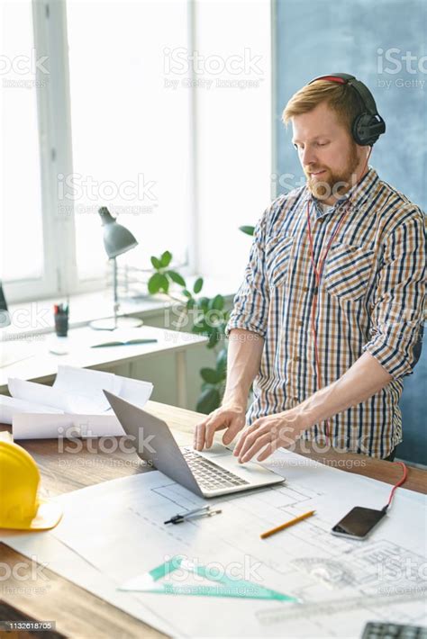 책상에 서 있고 프로젝트에서 작업 하는 동안 노트북에 디자인 프로그램을 사용 하는 헤드폰의 콘텐츠 잘생긴 엔지니어 건설 산업에