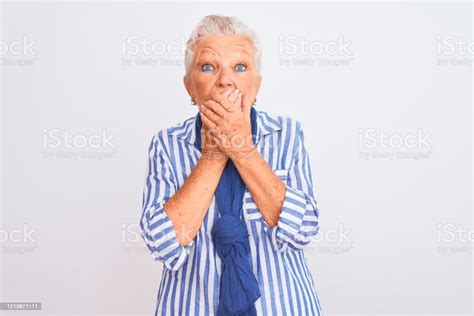 고립 된 흰색 배경 위에 서있는 파란색 줄무늬 셔츠를 입은 선임 회색 머리 여성은 실수로 손으로 입을 덮는 충격을 받았습니다