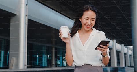 패션 사무복을 입은 성공적인 젊은 아시아 사업가는 뜨거운 음료의 일회용 종이컵을 들고 도시 현대 도시의 야외에 서 있는 동안 스마트폰을 사용합니다 이동 중 비즈니스 개념