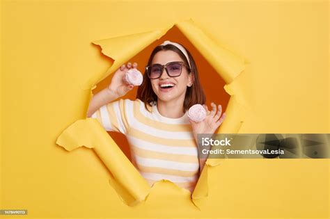 머리띠와 선글라스를 끼고 노란 종이 구멍에 서고 마시멜로를 들고 행복하고 맛있는 단 과자를 먹는 쾌활한 젊은 여성의 실내 사진 구멍에 대한 스톡 사진 및 기타 이미지