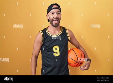 Middle Age Bald Man Holding Basketball Ball Over Yellow Background Sticking Tongue Out Happy