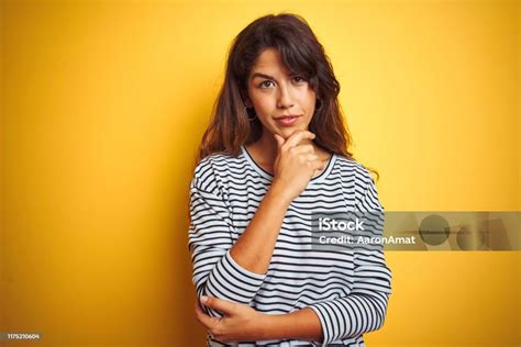 줄무늬 티셔츠를 입은 젊은 아름다운 여성은 턱에 손을 올려 놓고 미소를 지으며 카메라를 바라보며 고립된 배경을 바라보고 있습니다 긍정적 인 생각 T 셔츠에 대한 스톡 사진
