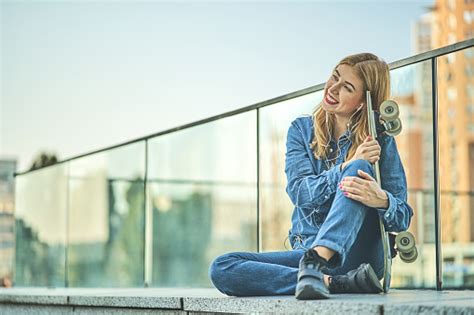 여름날 길거리 계단에 앉아 스케이트보드를 들고 있는 음악을 듣는 아름다운 세련된 미소 짓는 여성의 이미지 거리에 대한 스톡 사진