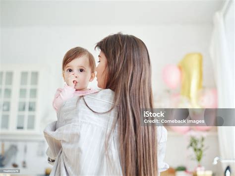 젊은 어머니의 뒷 전망 긴 머리 지주 아기 딸을 빠는 엄지 손가락과 얘기 그녀에 게 첫 번째 생일 엄지 손가락 빨기에 대한 스톡