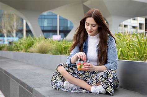 플라스틱 용기에서 과일을 먹고 콘크리트 좌석에 다리를 꼬고 뒤에 식물이있는 젊은 여성은 꽃 무늬 스커트 흰색 점퍼와 데님 재킷을 입고 있습니다 25 29세에 대한 스톡 사진