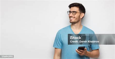 파란색 티셔츠와 안경을 쓰고 스마트폰을 손에 들고 서서 회색 배경에 고립된 공간을 복사하고자 하는 젊은 잘생긴 남자의 수평 배너