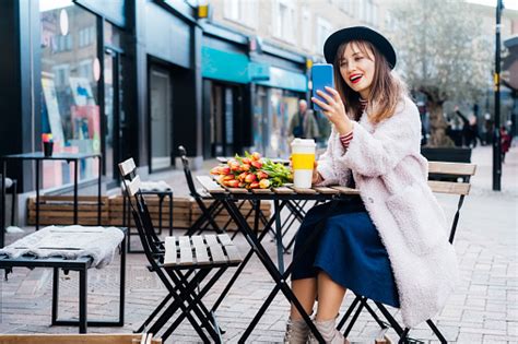 재사용 가능한 컵으로 커피를 마시고 길거리 카페에서 전화로 영상 통화를 하는 스타일리시한 젊은 웃는 여성 테이블에 신선한 튤립 꽃다발입니다 봄의 긍정적인 분위기 스트리트