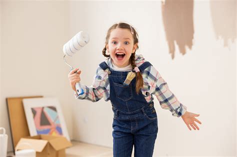 행복한 아이는 오른손에 페인트 롤러를 들고 방에서 수리 할 준비가되었습니다 소녀는 데님 작업복과 체크 무늬 셔츠를 입고 있습니다 배경에는 벽과 수리를위한 물건이 그려져
