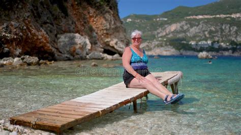 Elderly Senior Woman In Swimsuit Resting On Small Wooden Boat Pier Near Calm Clear Sea Beach