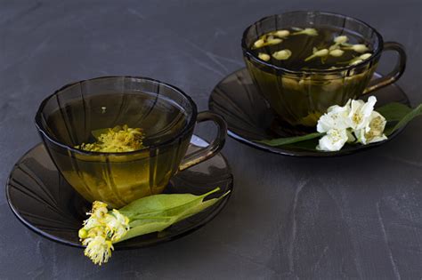 꽃과 차 한 잔 검은 색 배경에 고립 된 꽃꽃이있는 허브 핫 린든 차와 재스민 차가있는 두 개의 어두운 유리 컵 테이블에 녹색