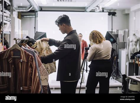 Side View Of Male Designer Choosing Clothes From Rack During Photo