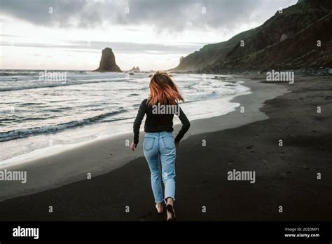 Back View Of Barefoot Woman In Casual Outfit Running On Wet Sand Near