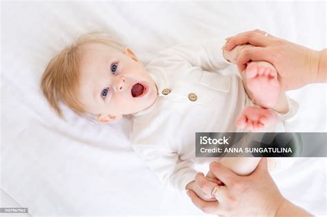 6 개월 된 아기의 클로즈업 초상화 파란 눈을 가진 금발 소년이 흰색 면화 바디 수트를 입은 밝은 침실의 흰색 침대에서 하품
