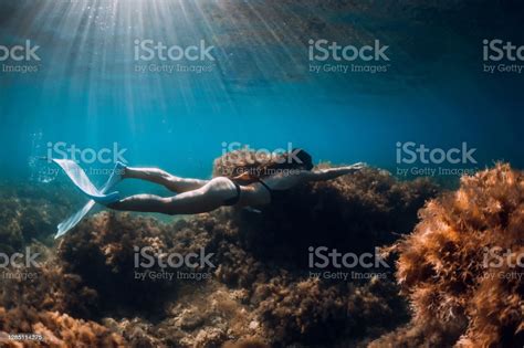 하얀 지느러미를 가진 프리다이버 소녀가 해초와 태양광선으로 바위 근처에서 활공합니다 푸른 바다에서 프리 다이빙과 아름다운 빛