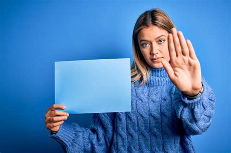 진지하고 자신감있는 표정 방어 제스처로 정지 표지판을하고 열린 손으로 고립 된 파란색 배경 위에 서있는 배너를 들고있는 젊은 아름다운 여성 갈색 머리에 대한 스톡 사진 및