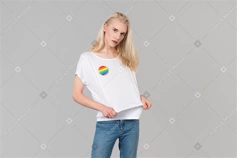 긴 금발 머리를 가진 잘 생긴 청년 회색 배경에 서서 청바지와 Lgbt 배지가있는 흰색 티셔츠를 입고 사진