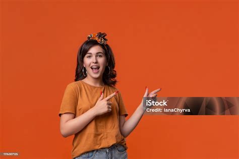 티셔츠와 오렌지 배경 위에 고립 된 권리 측에서 그녀의 손가락으로 가리키는 아름다운 머리띠에 예쁜 갈색 머리 여자 광고를 위한 장소입니다 T 셔츠에 대한 스톡 사진 및 기타