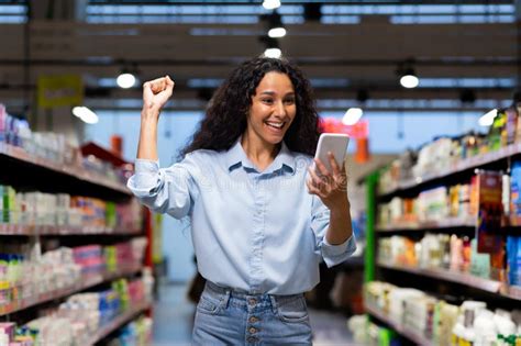 Excited Young Woman Celebrating Success With Smartphone In Supermarket