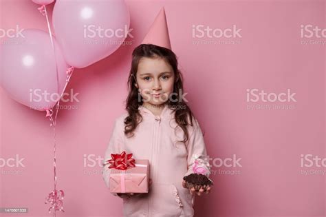 분홍색 옷과 파티 모자를 쓴 행복한 어린 소녀 생일을 위한 귀여운 선물과 촛불이 든 컵케이크를 들고 있다 광고에 대한 스톡 사진