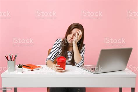 젊은 여자에 기대어 자 고 피곤된 하면서 차 또는 커피 한잔 들고 작업과 파스텔 핑크 배경에 고립 된 Pc 노트북으로 사무실에