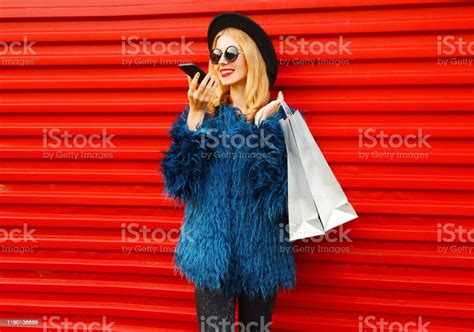 음성 명령 레코더를 사용하여 스마트 폰을 들고 쇼핑 백을 가진 세련된 젊은 여성 보조 또는 빨간 벽 배경 위에 파란색 인조 모피 코트 검은 색 둥근 모자와 선글라스를 입고