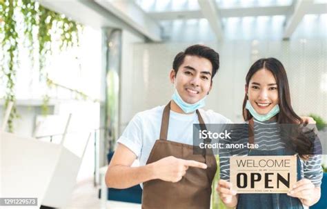 앞치마에 매달려 있는 비즈니스 오너 매력적인 젊은 아시아 커플우리는 새로운 카페에 고객을 환영하는 사인을 엽니다 카페에 서있는 동안 열린 표지판을 들고 보호 얼굴 마스크와