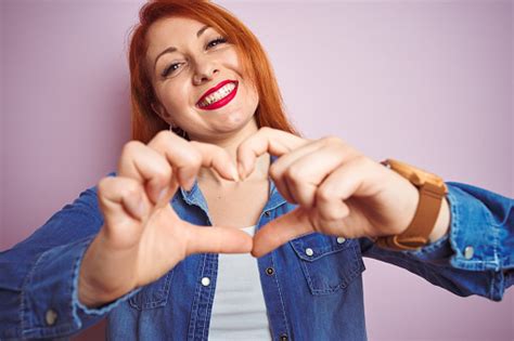 Youg 아름다운 빨간 머리 여자는 손으로 심장 기호와 모양을 보여주는 사랑에 미소 고립 된 분홍색 배경 위에 서 데님 셔츠를