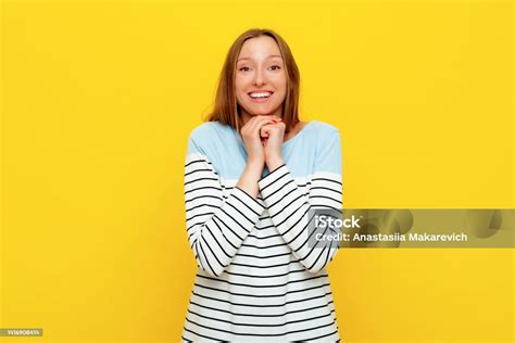 턱 밑에서 손을 잡고있는 귀여운 페어 헤어 여성은 놀랍고 행복한 표정을 짓고 흥분하고 긍정적 인 감정을 표현하고 노란색 배경 위에 줄무늬 스웨트 셔츠를 입고 서 있습니다