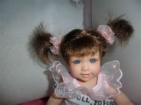 A Doll With Blue Eyes And Brown Hair Wearing A Pink Dress Sitting On A Bed