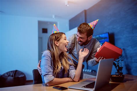 젊은 깜짝 놀라게 사랑 스러운 백인 갈색 머리 와 생일 모자 머리에 식탁에 앉아 생일 선물을 받고 그녀의 남자 친구는 그녀에게 선물을주고 식탁에는 노트북과 스마트폰이