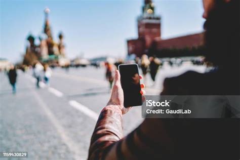 빨간 광장에서 걷는 동안 빈 화면으로 휴대 전화를 들고 있는 힙스터 남자 광고를위한 모형 영역이있는 스마트 폰 장치에서 웹 카메라를 통해 셀카 사진을 만드는 남성 관광객의