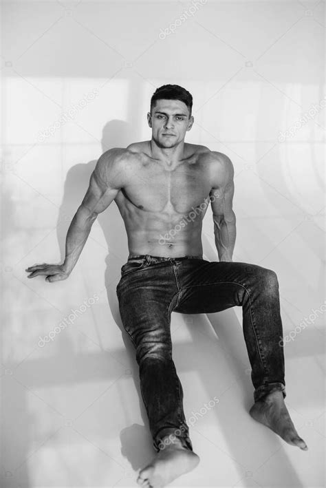 Un joven atleta sexy con abdominales perfectos está sentado en el estudio en topless en jeans