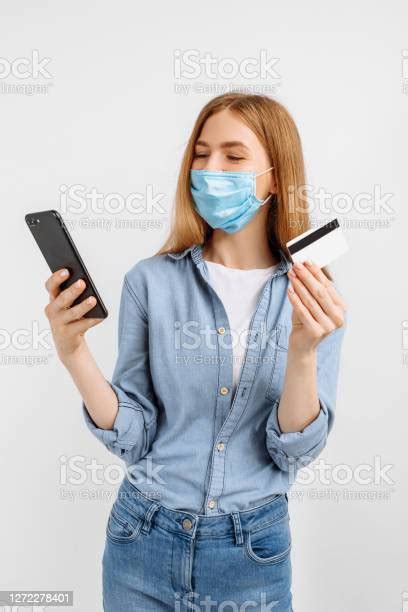 얼굴에 의료 용 마스크를 쓰고 플라스틱 신용 카드를 보여주고 흰색 배경에 휴대 전화를 사용하는 젊은 여성 격리에 대한 스톡 사진