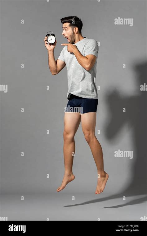 Shocked Caucasian Man With Alarm Clock In Hand Jumping After Waking Up