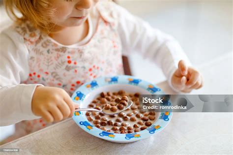 아침 식사로 우유와 함께 건강한 시리얼을 먹는 유아 소녀의 닫습니다 부엌에 앉아 귀리 시리얼을 준비와 재미 다채로운 옷에 귀여운 행복 아기 아이 집 안의 실내 가정 생활에