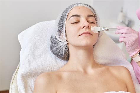 그녀의 피부에 얼굴 마스크를 적용 하는 동안 미용사는 동안 Couh에 누워 매력적인 여자 뷰티 살롱에서 화장품 치료를 즐기는 아름 다운 Cherful 여성 피부과 스킨케어