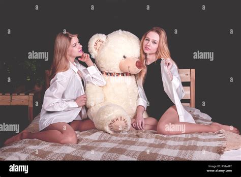 Two Cute Girlfriends In White Shirts Are Sitting On The Bed With A Big