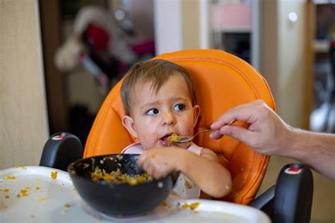 주황색 자식좌석에 앉은 귀여운 어린 소녀는 접시에 손을 넣고 부모가 들고 있는 숟가락에서 먹고 부모를 바라봅니다 클로즈업 전면 보기 소프트 포커스 블러 배경 건강한 식생활에